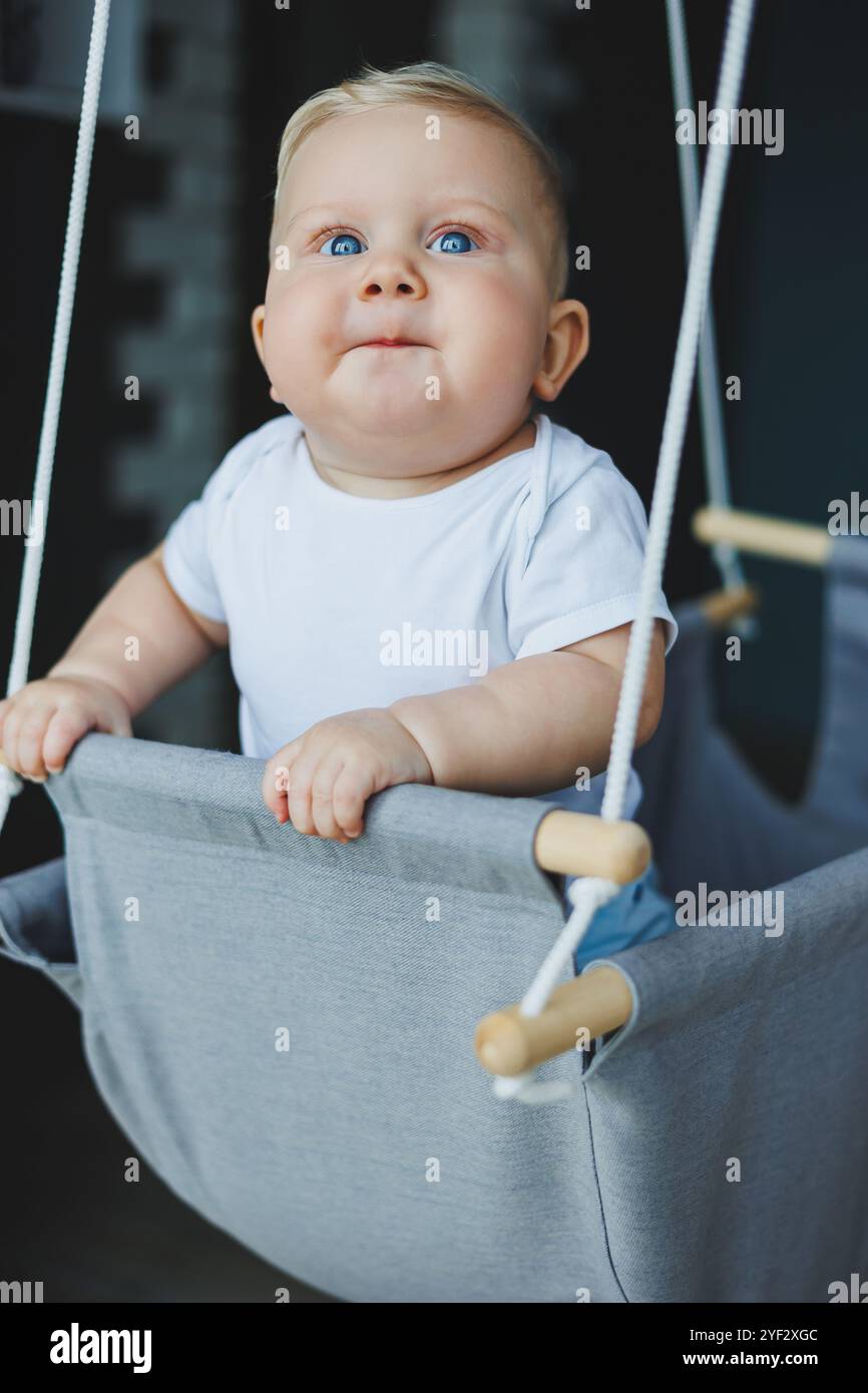 Baby boy swings in a swing at home. The child learns to have fun. Child ...