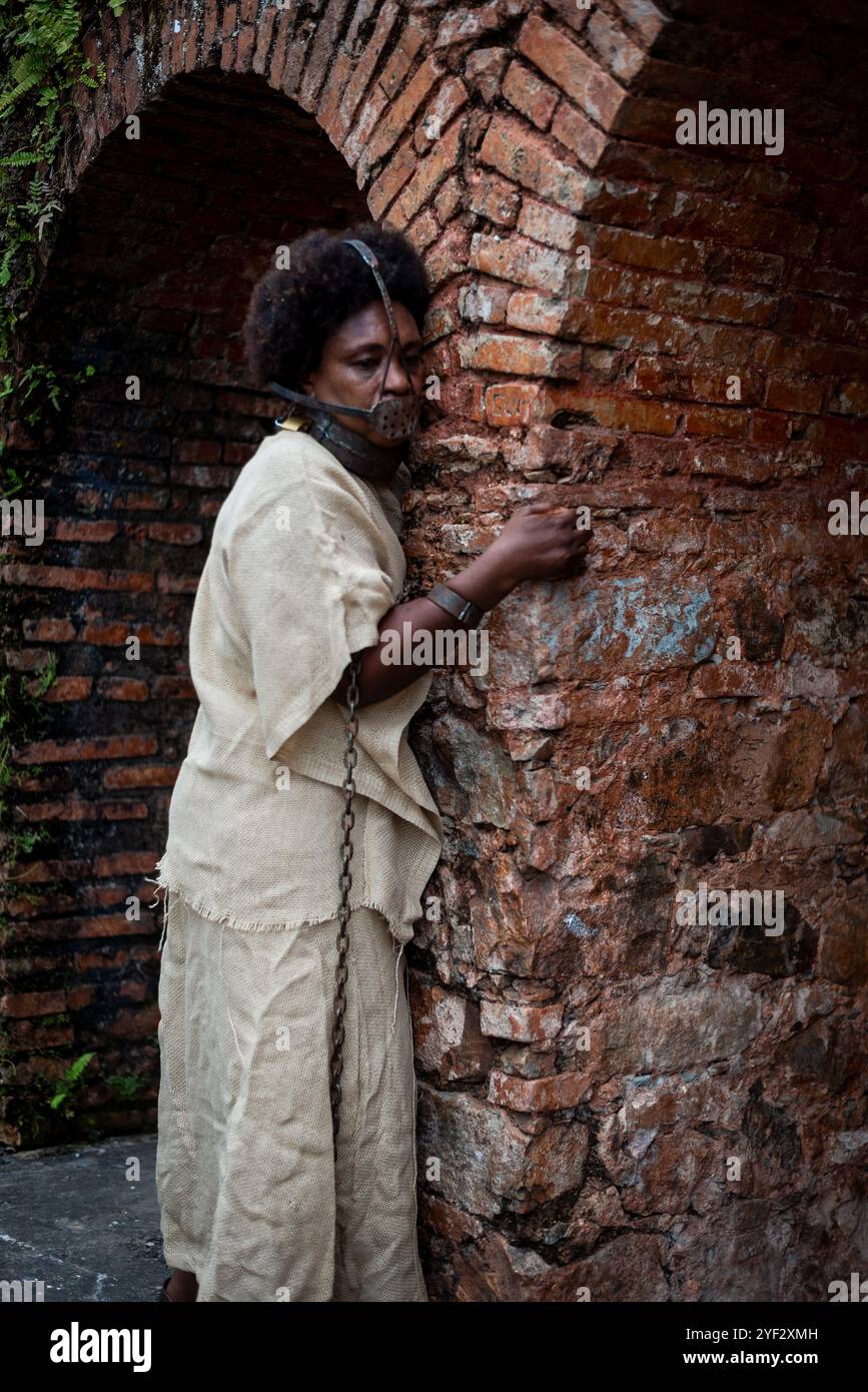 Black woman chained and with an iron mask stuck in her mouth, standing, leaning against a brick ...