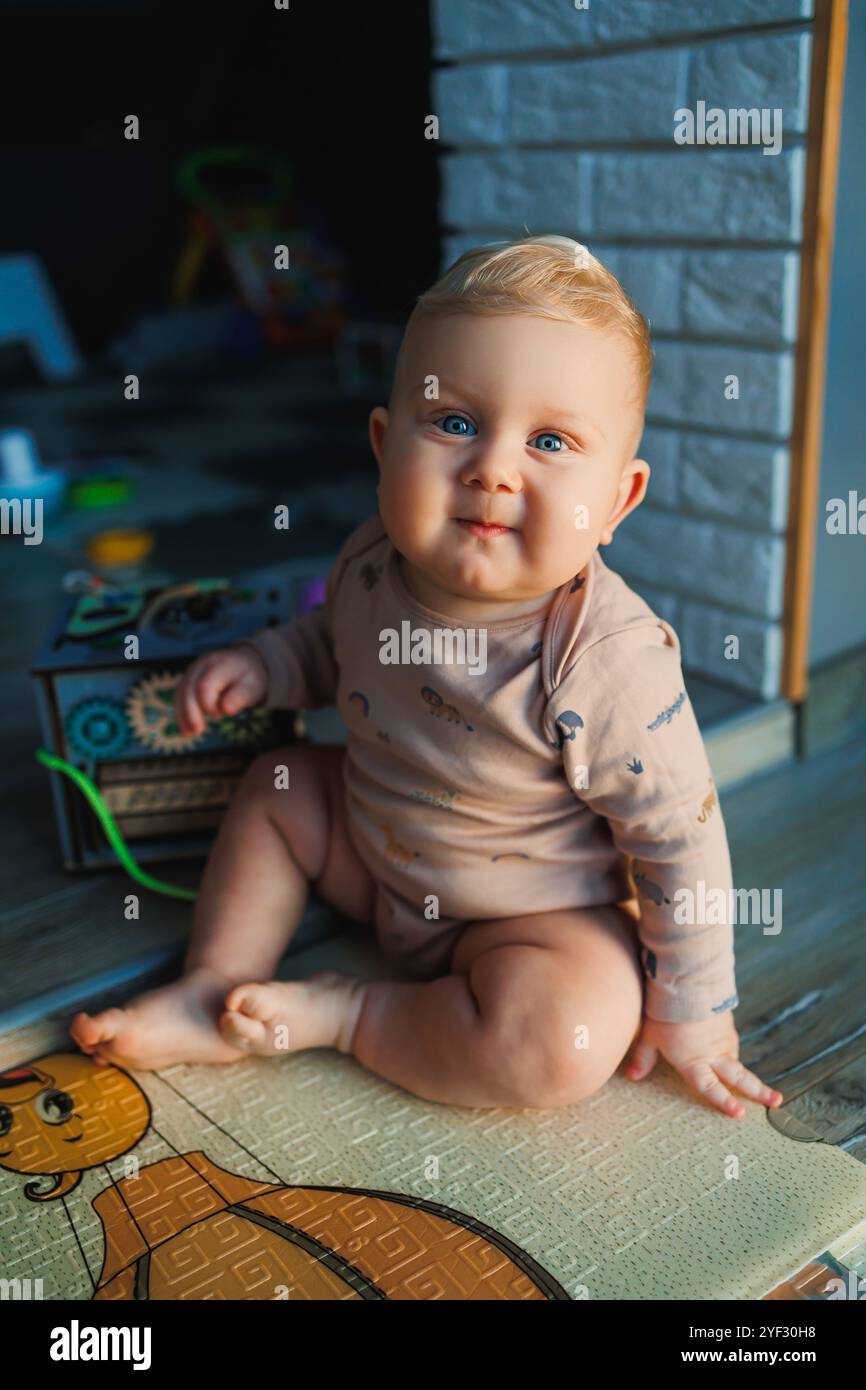 Fair-haired boy with blue eyes baby 11 months in a bodysuit sits on the ...
