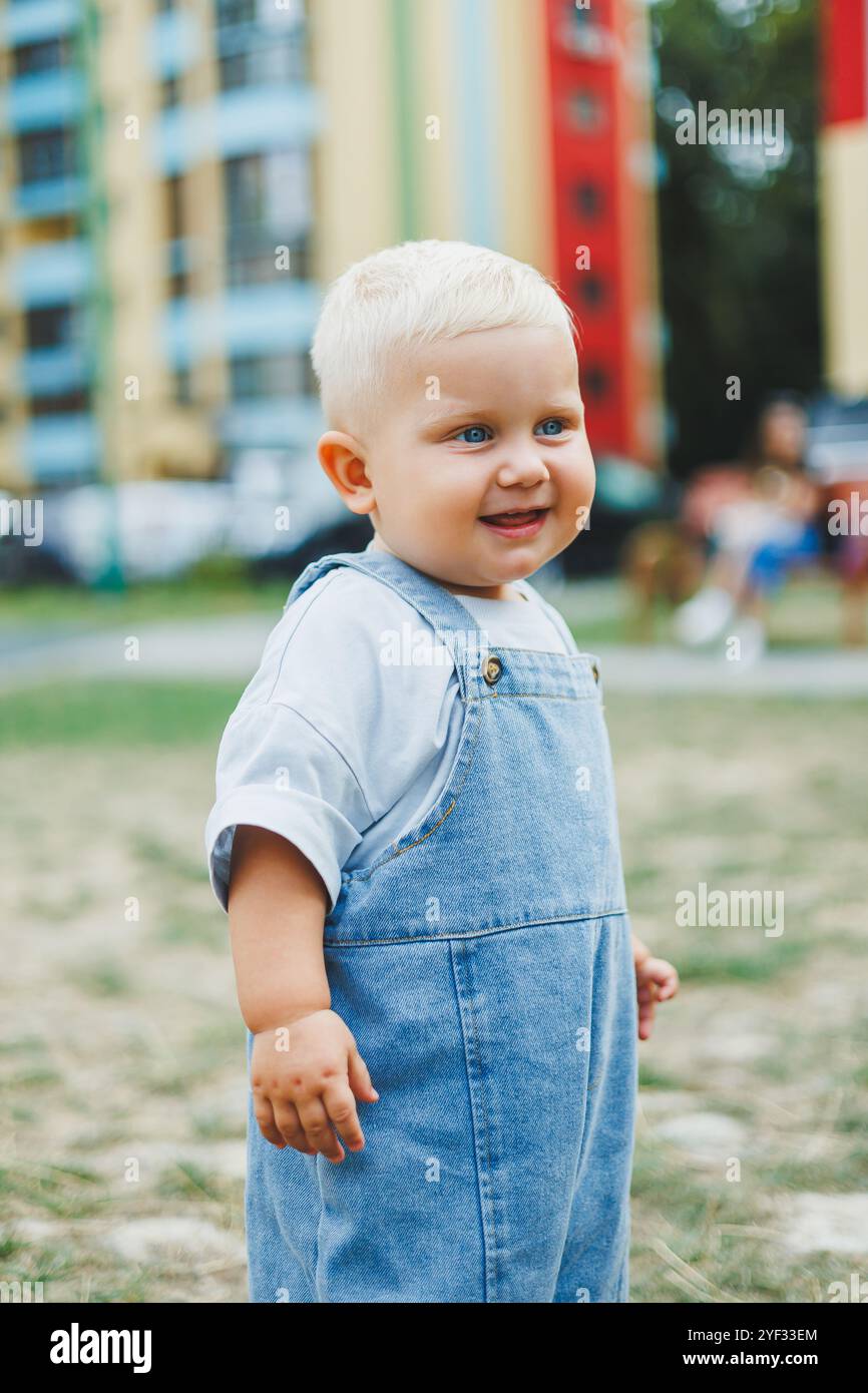 A 1-year-old baby boy in denim overalls is walking on the street ...