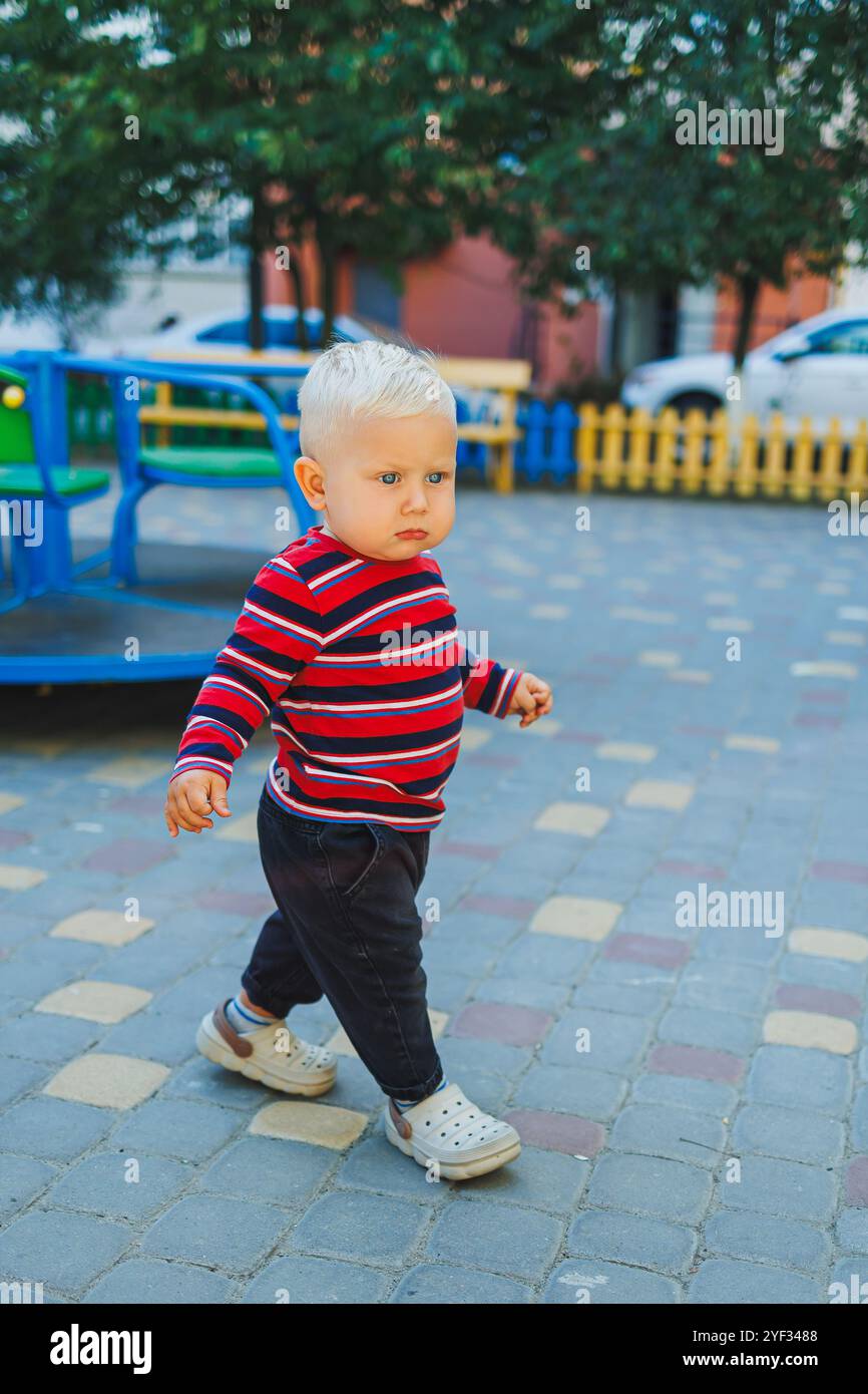 A little blond boy, 1 year old, walks on the children's playground. The ...