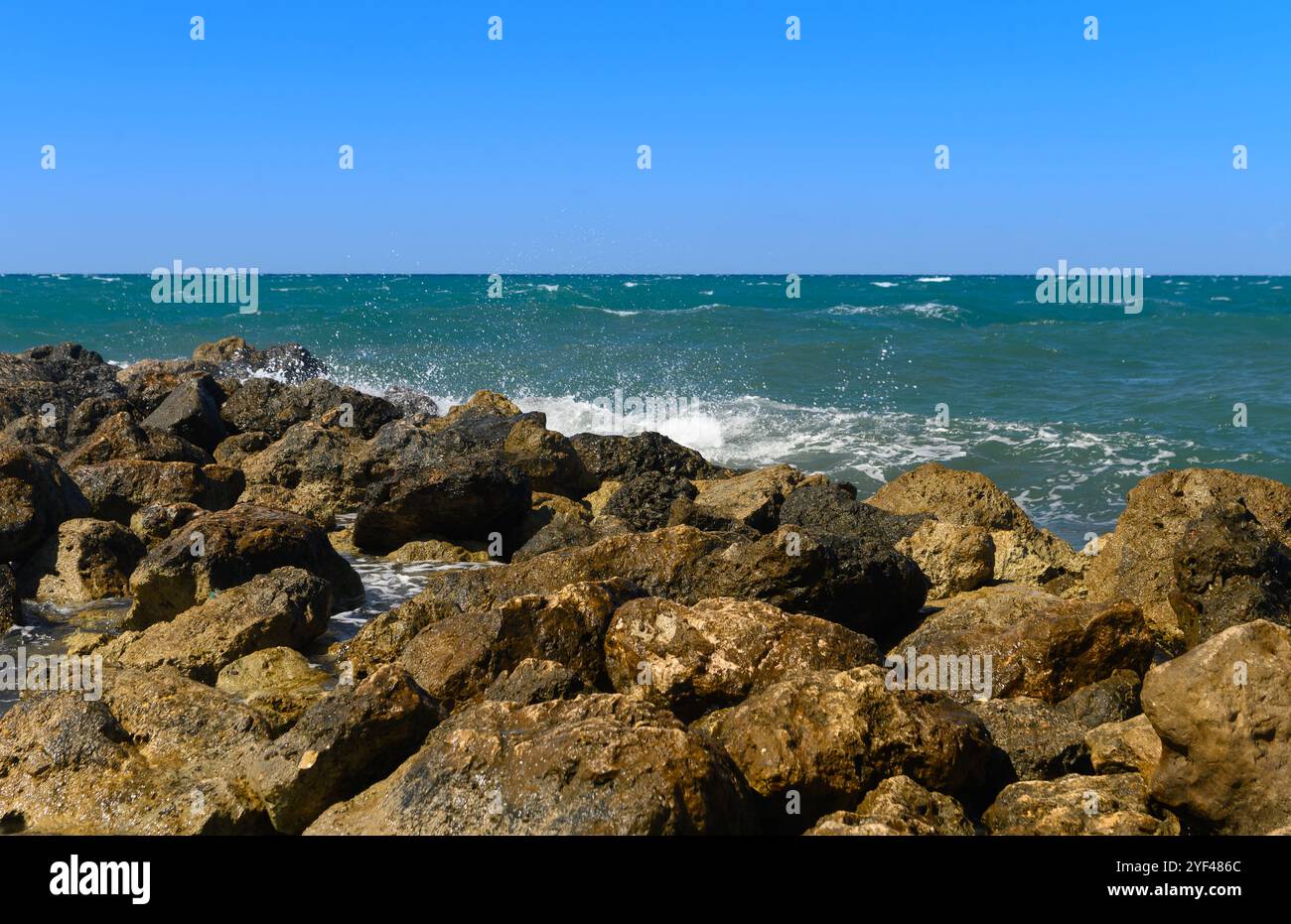 The sunlit coastline features waves splashing dramatically over a ...