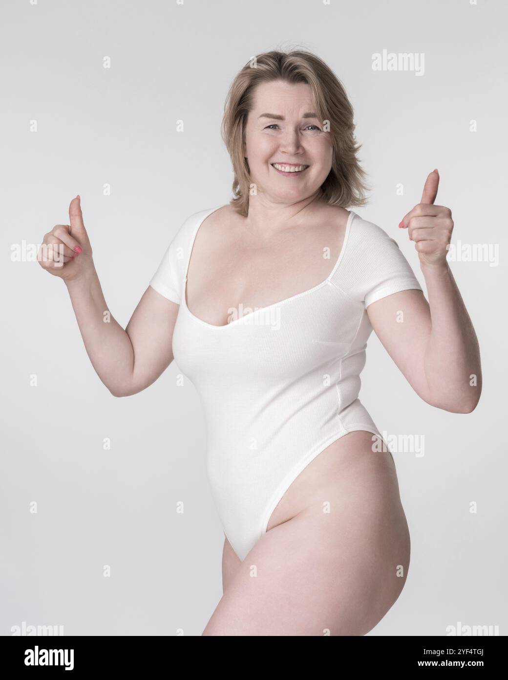 Portrait of plus size woman in bodysuit raising both hands and showing thumbs up, posing on ...