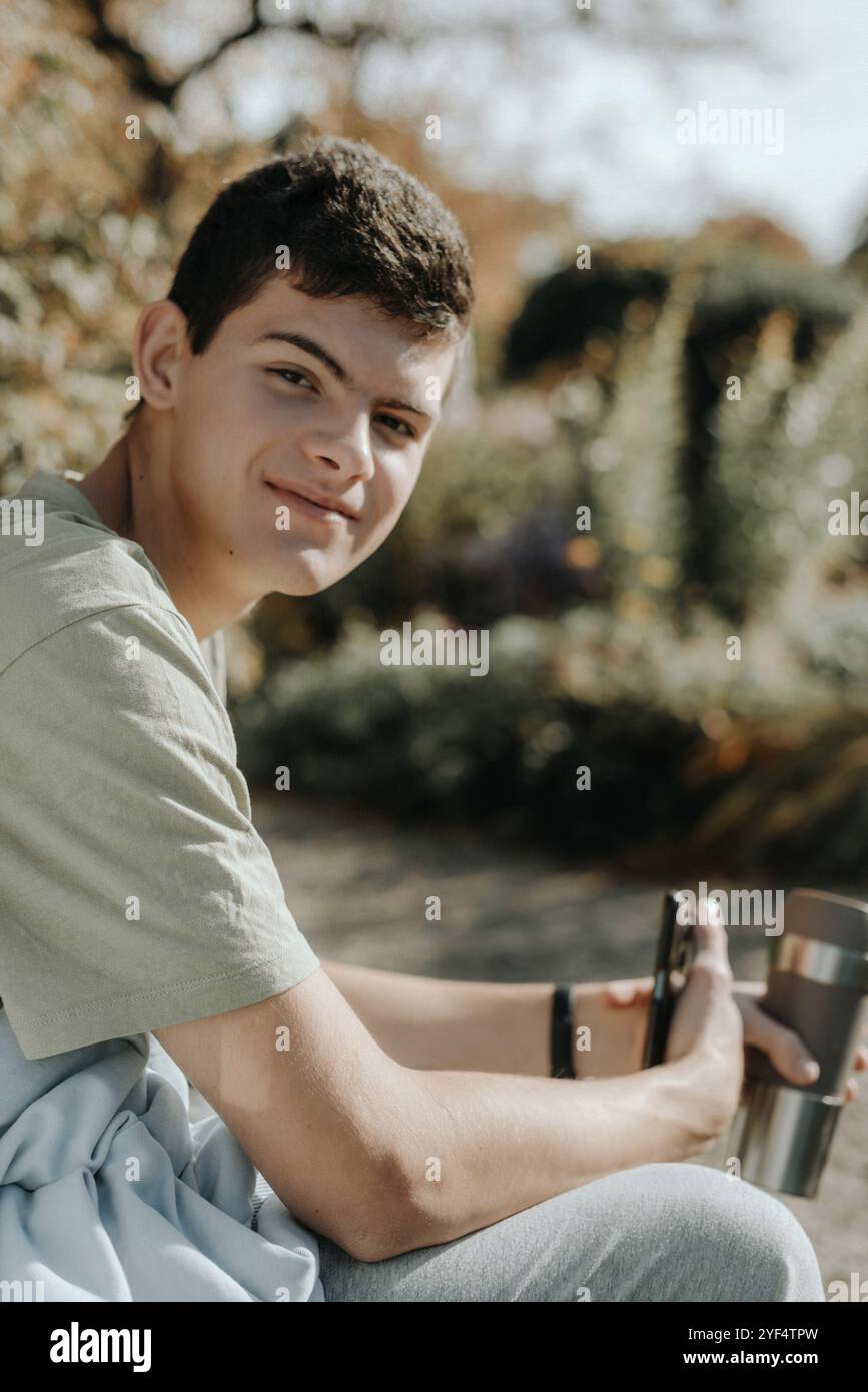 A teenager sits on a bench in the park drinks coffee from a thermo mug ...