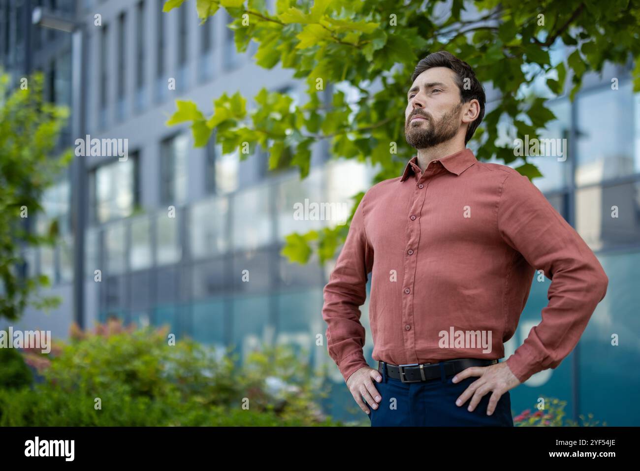 Mature man stands confidently outdoors, hands on hips, looking upwards ...