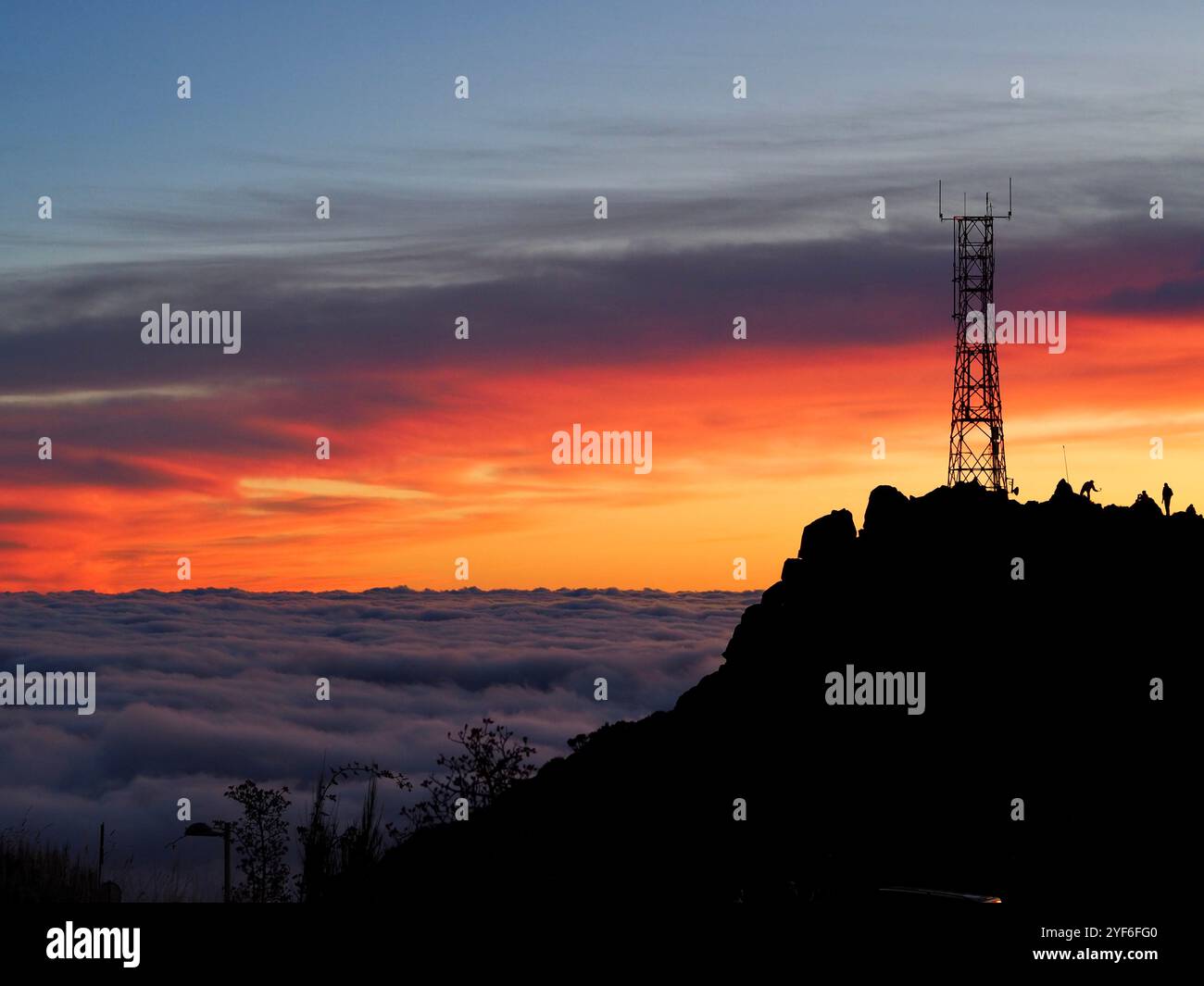 A tall cell tower stands silhouetted against a vivid sunset, its ...