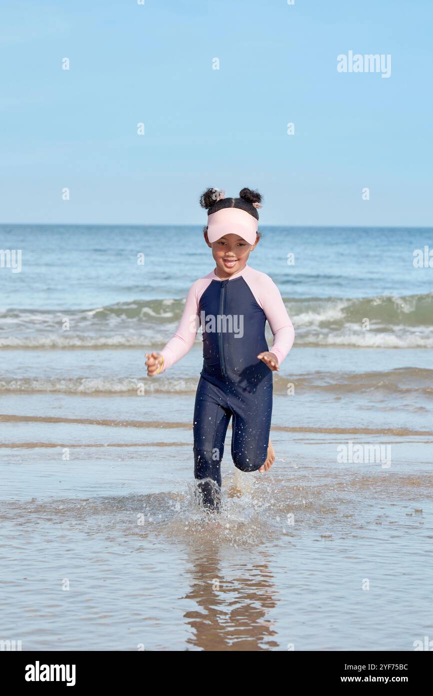 Smiling girl in a one piece protective surf suit and sun visor running ...