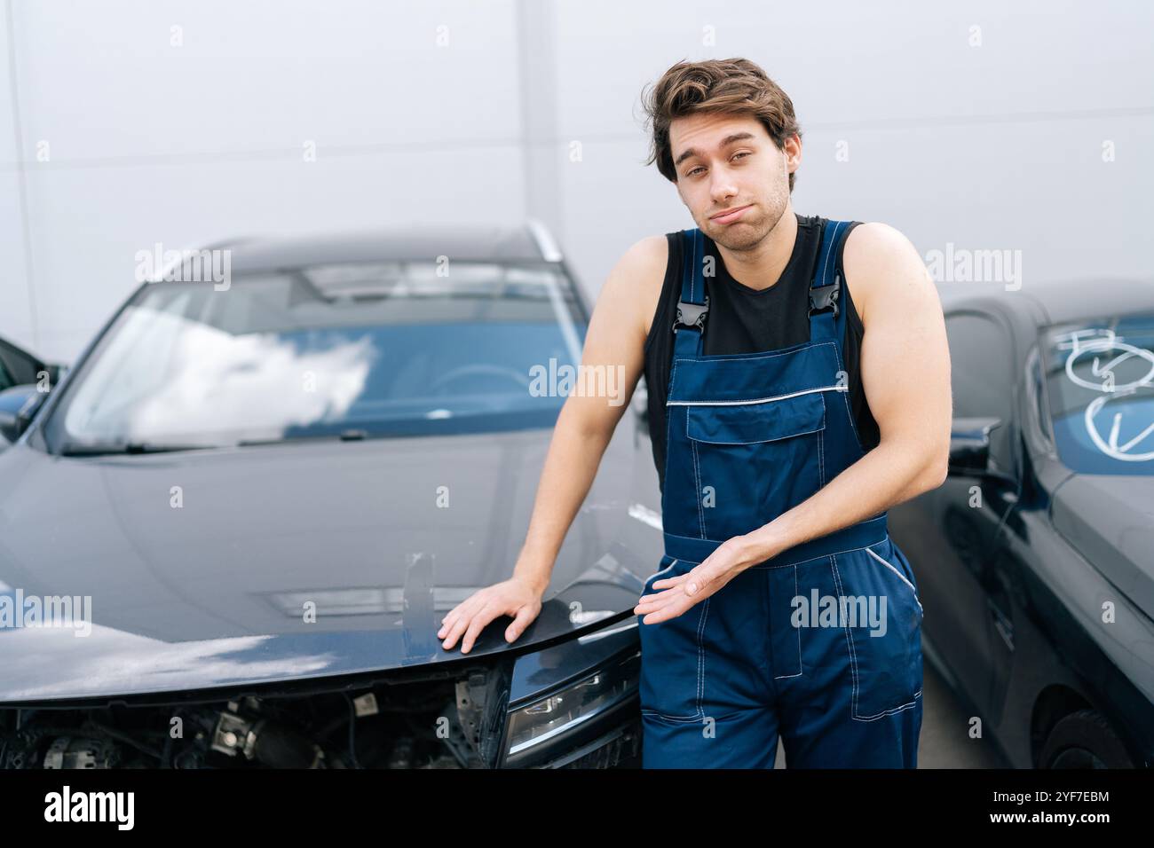 Puzzled mechanic male in blue overalls standing next to damaged car ...