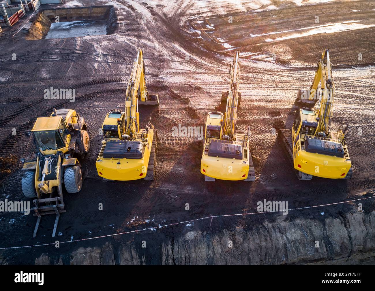 Großbaustelle - aufgereihte, gelbe Bagger parken neben dem Rohbau einer ...