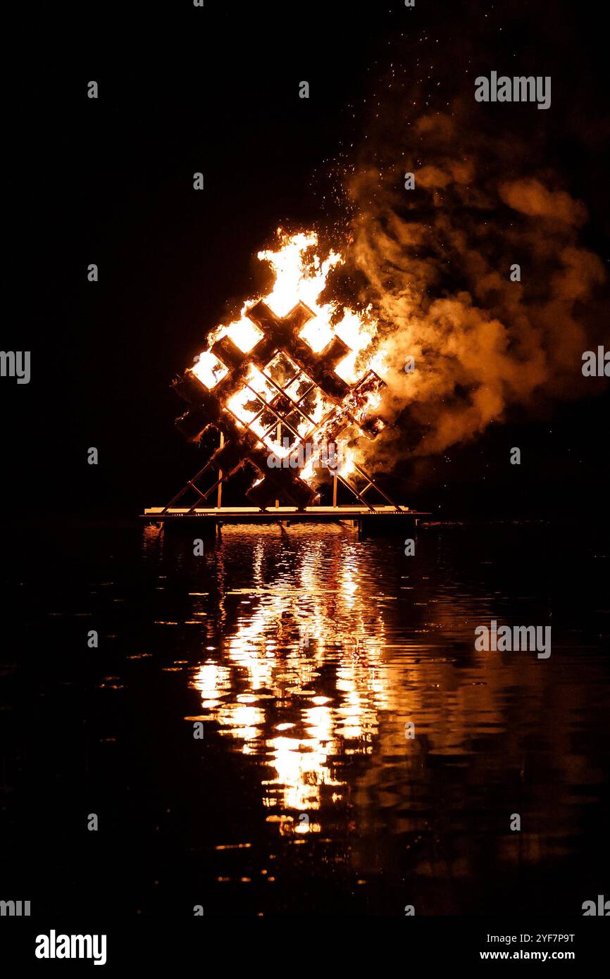 A large wooden lattice structure burns on a platform over water, with ...
