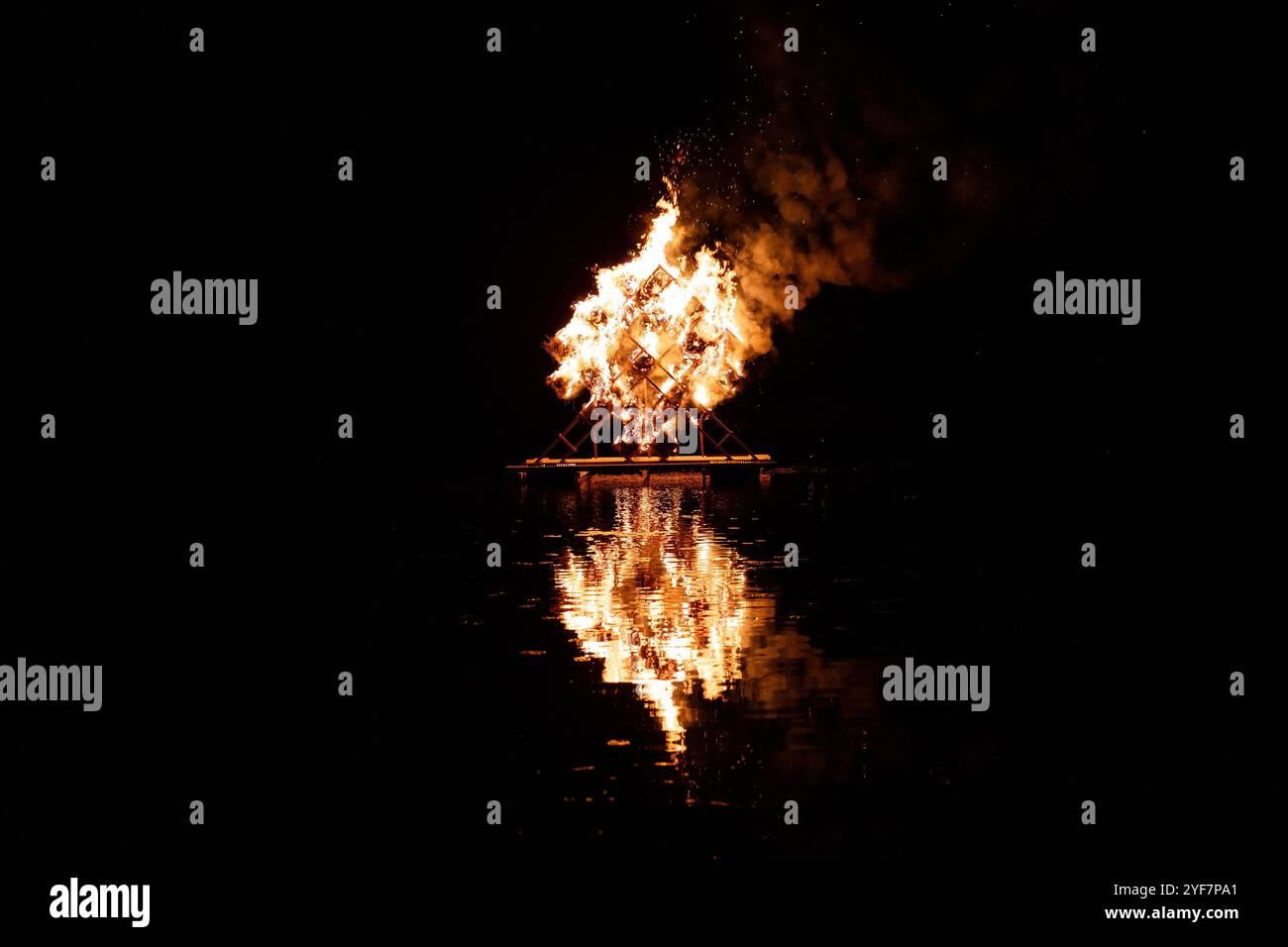 A large wooden structure is engulfed in flames, floating on a calm body ...