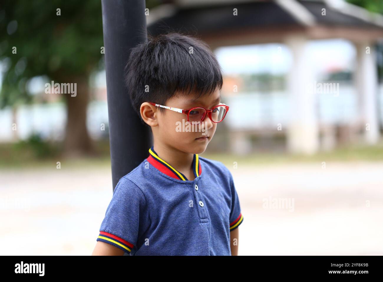 A young boy with glasses stands thoughtfully at a playground. His ...