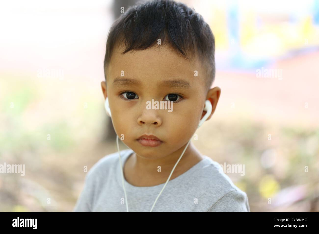 A young boy sits outdoors, wearing earbuds and listening intently. His ...