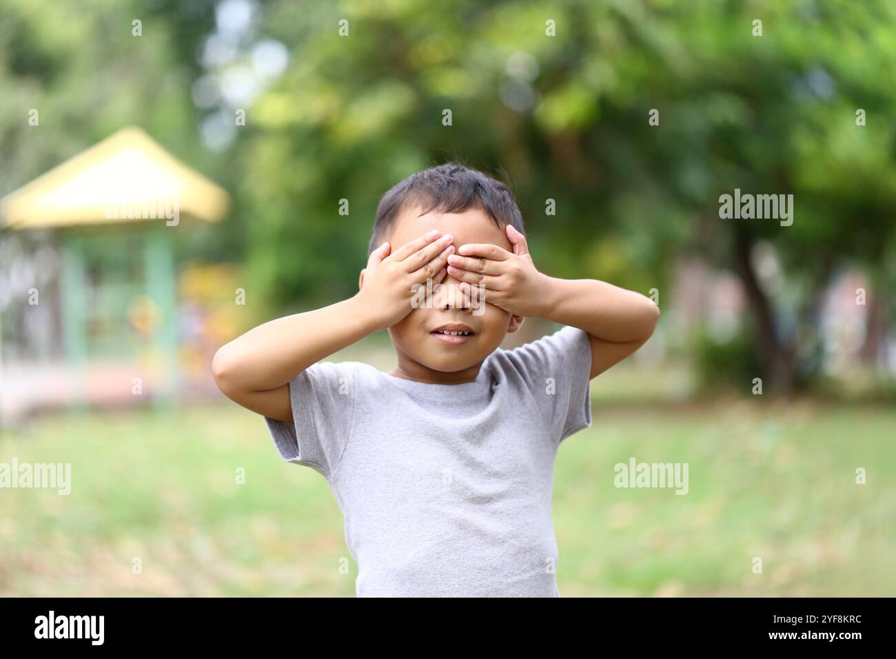 A joyful child plays hide and seek in a green park, covering his eyes ...