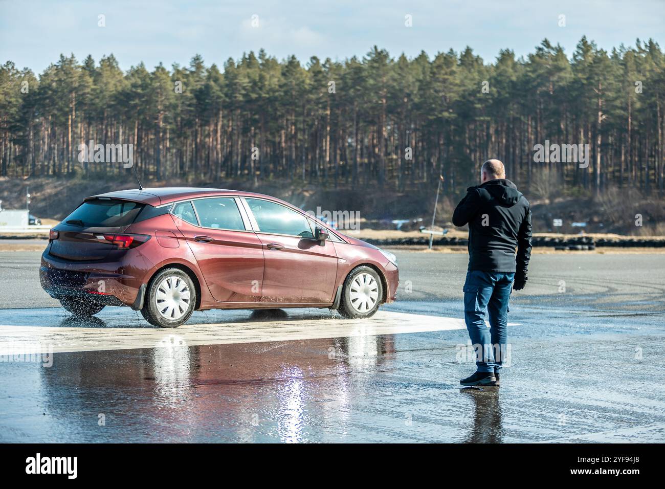 man observing a wet road test with a maroon car skidding on a water ...