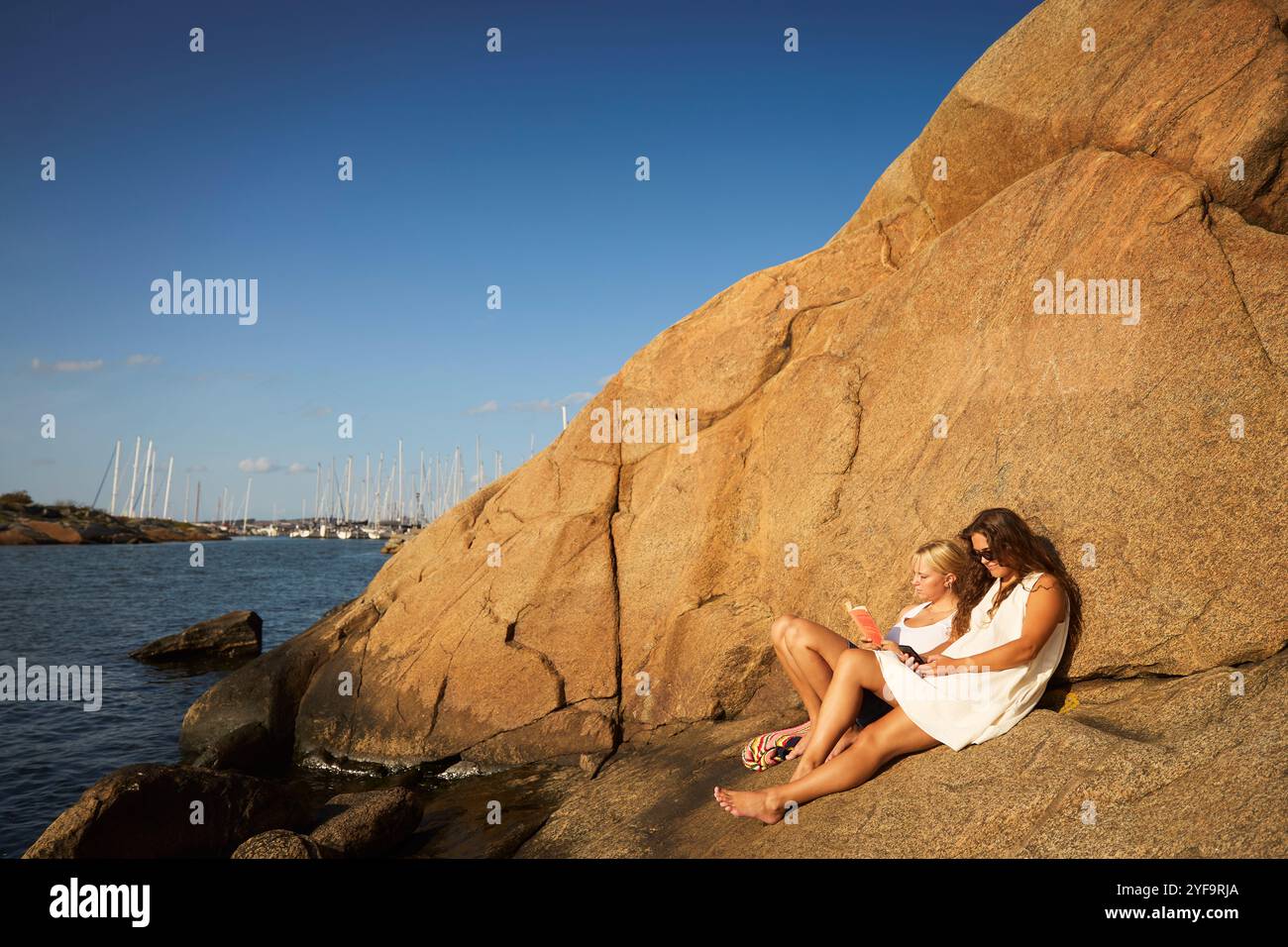 Mid adult woman reading book and female friend using mobile while ...