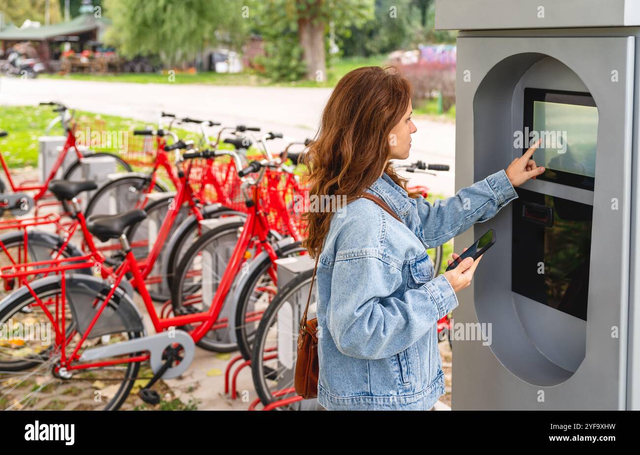 Urban woman interacting with the screen of a bike-sharing kiosk to pay and unlock a bicycle at a ...