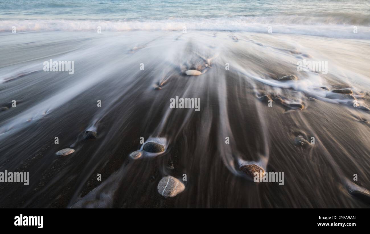 Pebbles in the beach on a black sand and flowing sea water creating ...