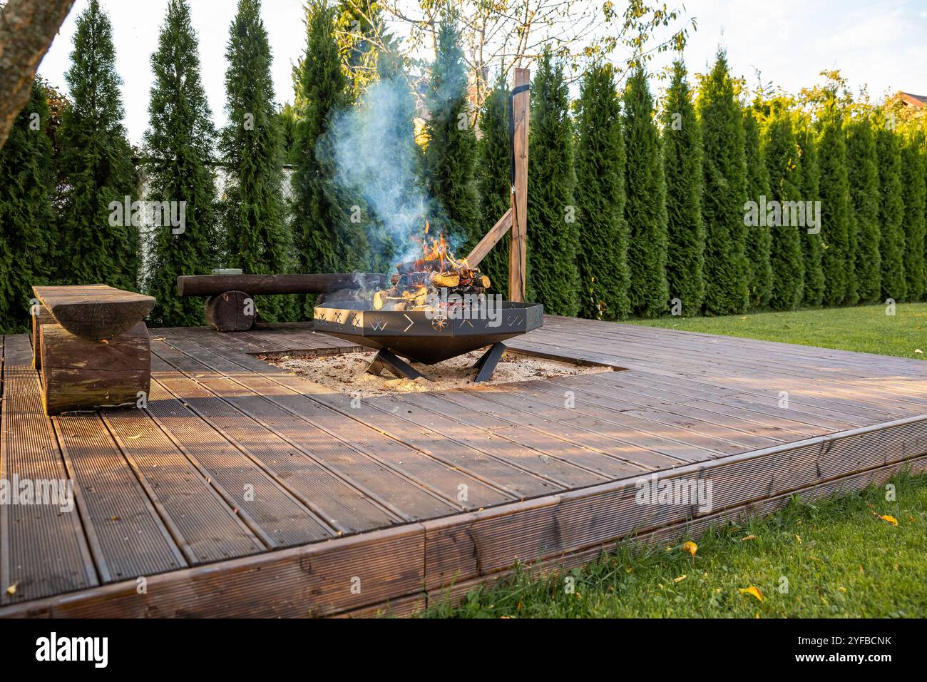 A wooden deck features a circular fire pit with burning logs and smoke ...
