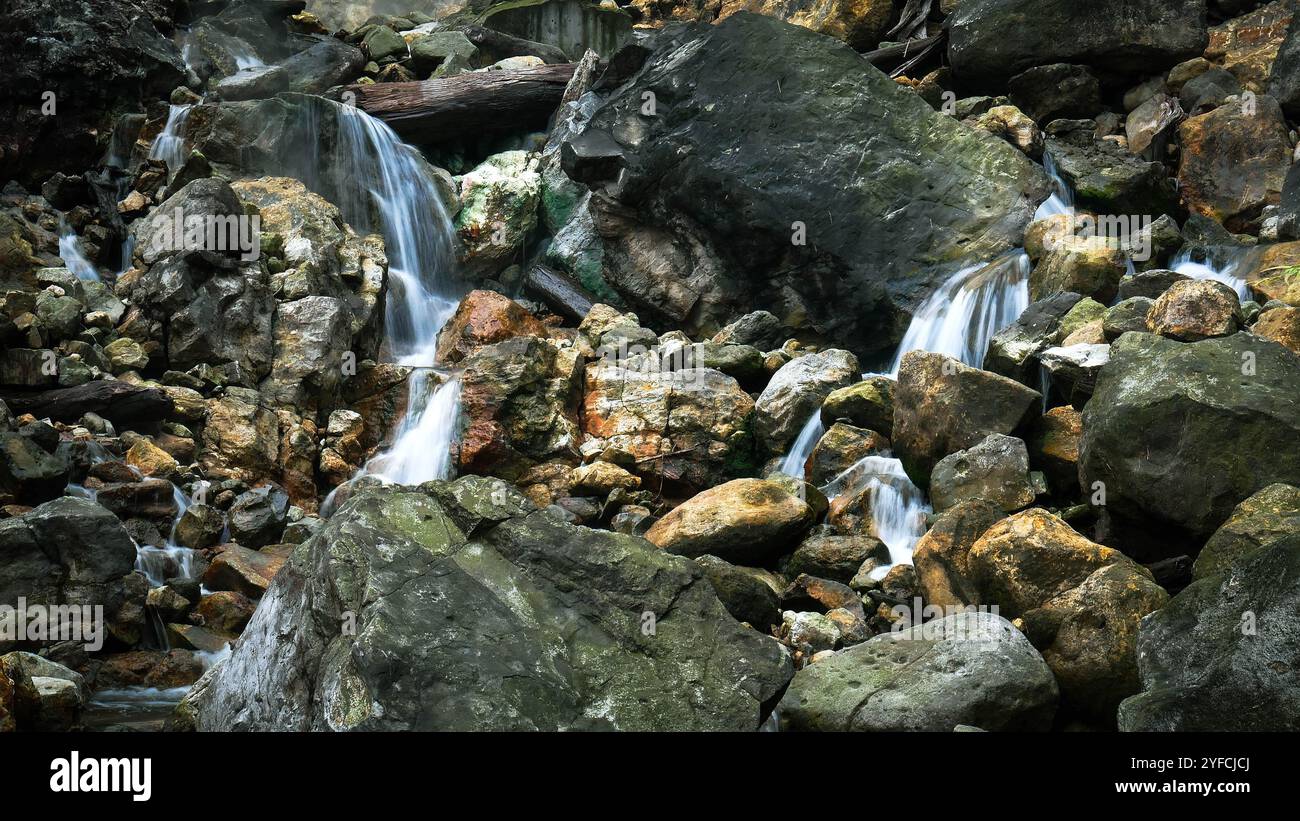 A mini waterfall with rushing water flows over large rocks covered in ...