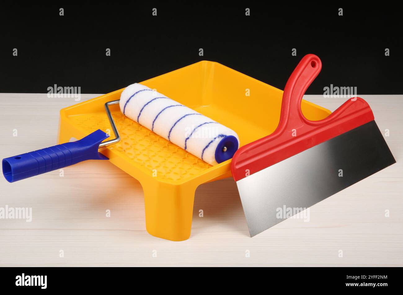 Paint roller, tray and putty knife on light wooden table against black background. Construction ...