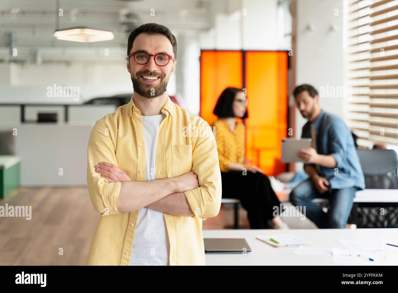 Young businessman is smiling with his arms folded in a busy office with ...