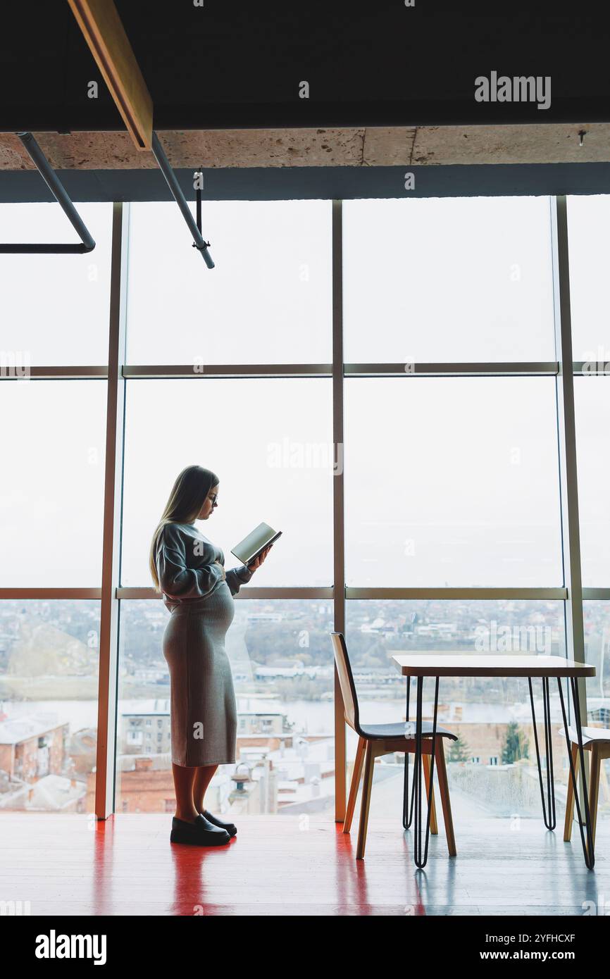 Young pregnant woman at work in the office standing near the window ...