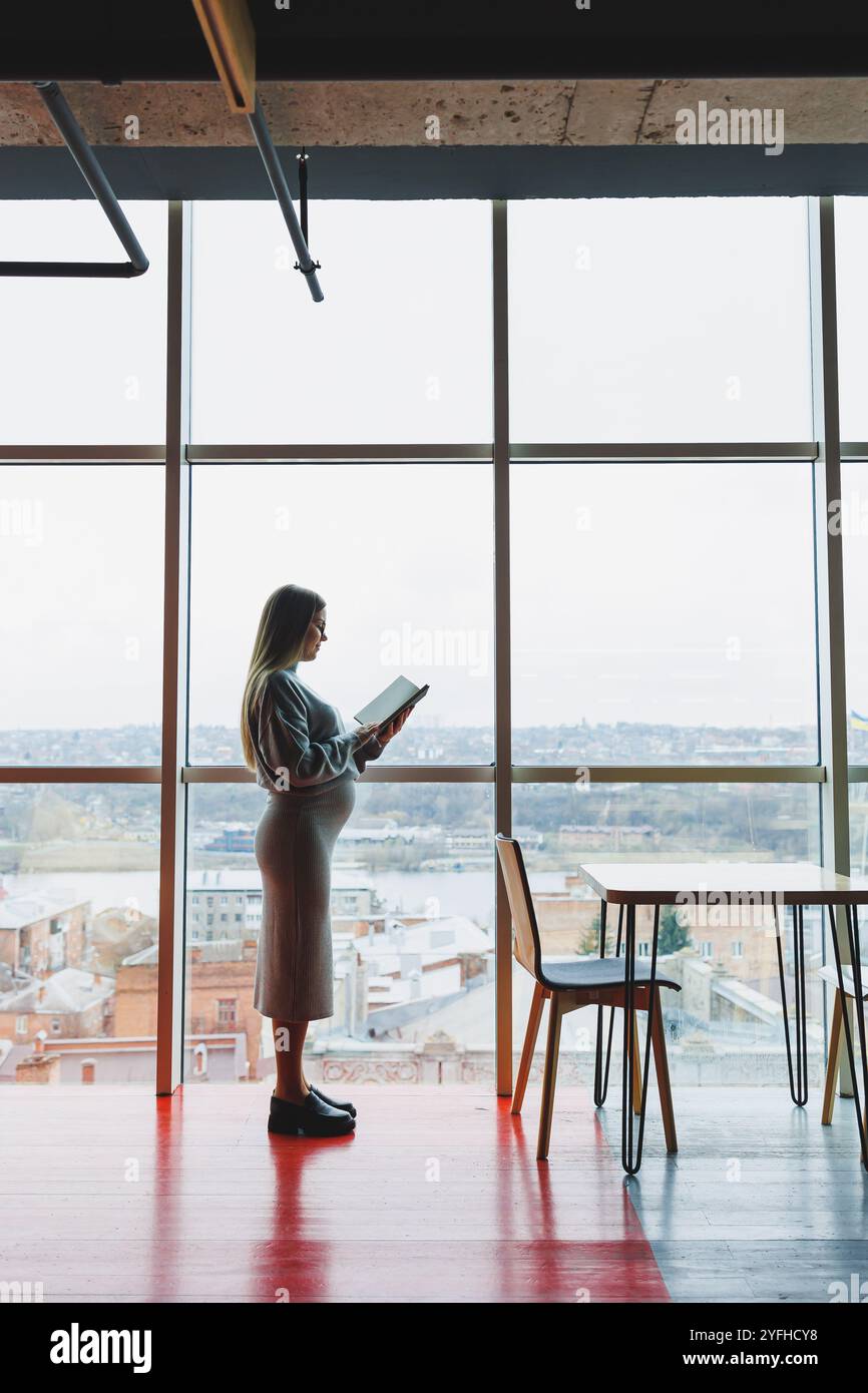 Young pregnant woman at work in the office standing near the window ...