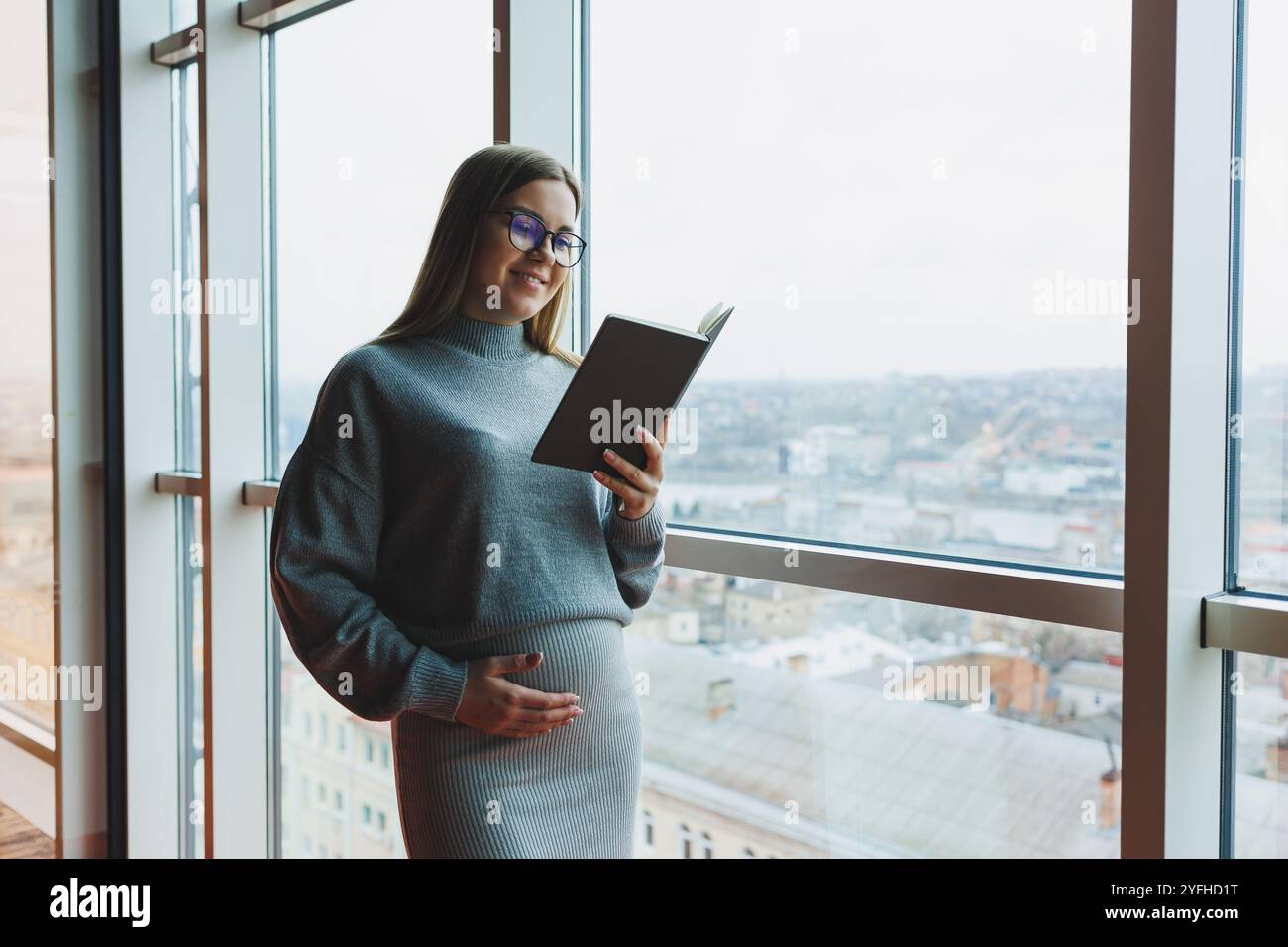 Young pregnant woman at work in the office standing near the window ...