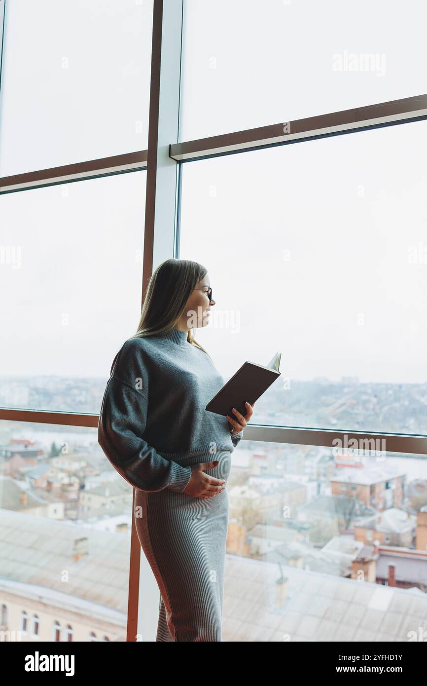 Young pregnant woman at work in the office standing near the window ...