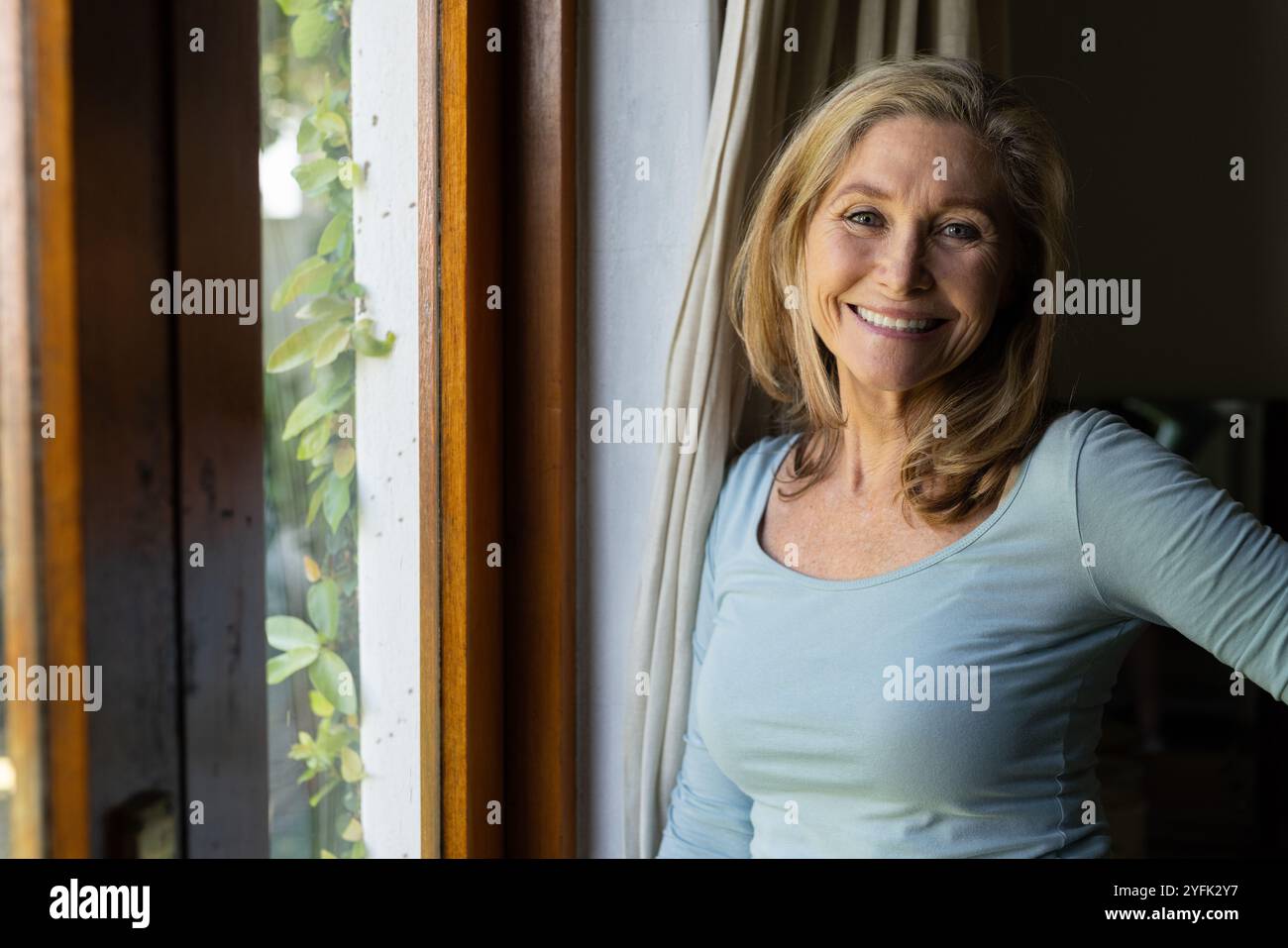 Smiling senior woman standing by window at home, enjoying natural light ...