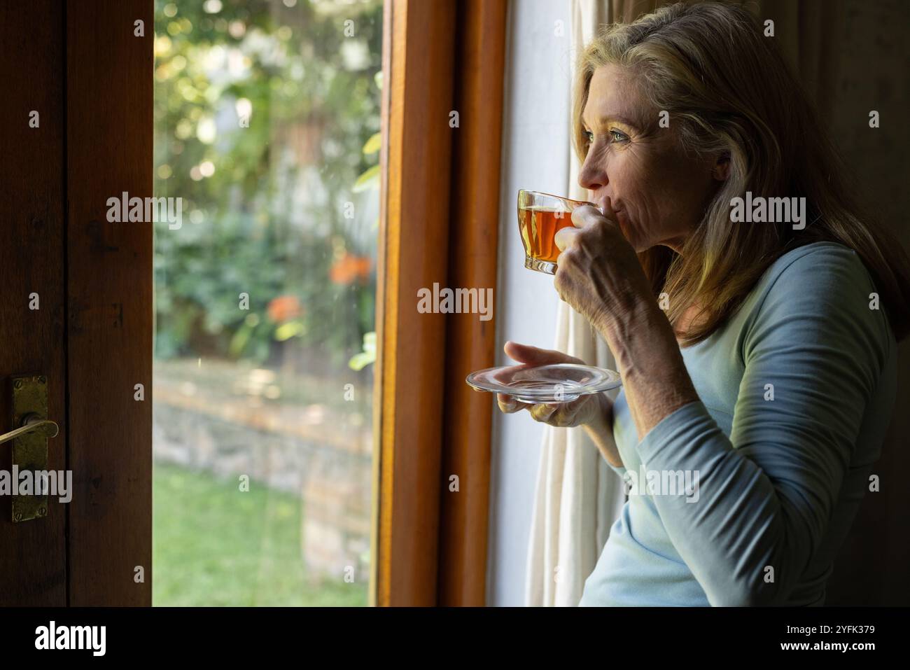 senior woman enjoying tea by window, peacefully gazing at garden view ...