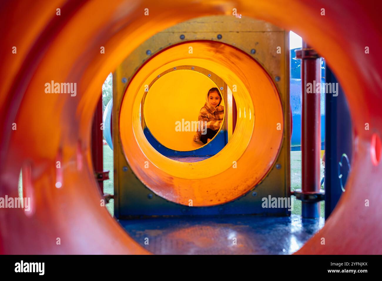 A child sits inside a vibrant orange playground tunnel, seen through circular openings, creating ...