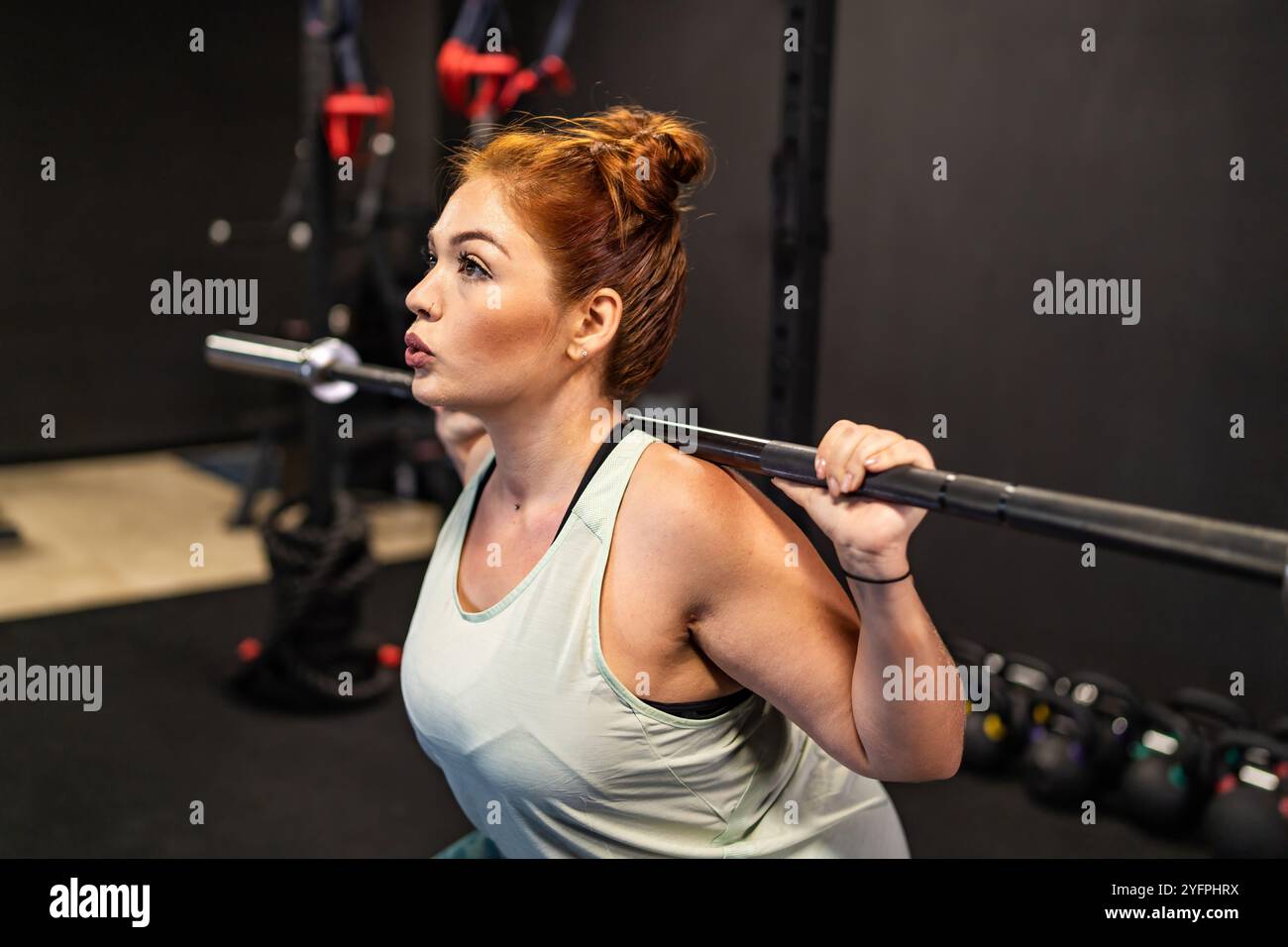 Focused redhead woman performing barbell squat exercise in modern dark ...