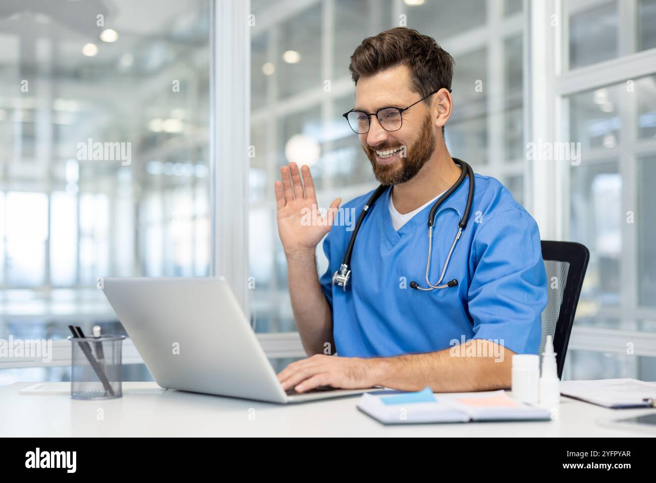 Smiling doctor in blue scrubs participates in virtual consultation ...