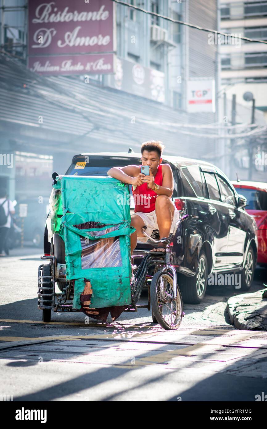 A young filipino man takes a break sat on his trike and checks his ...
