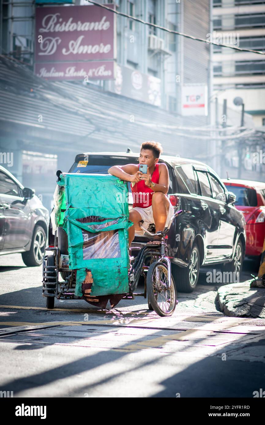 A young filipino man takes a break sat on his trike and checks his ...