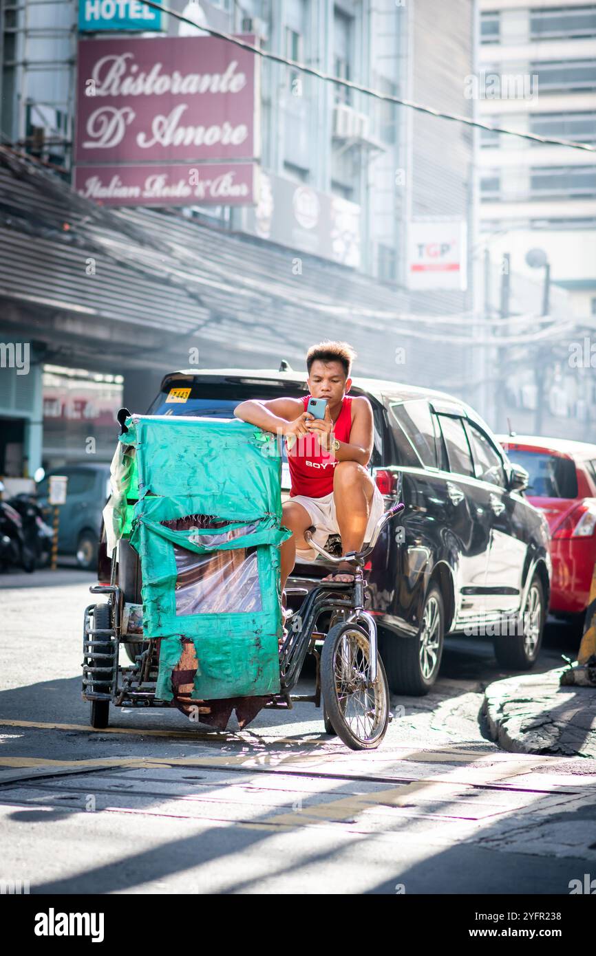 A young filipino man takes a break sat on his trike and checks his ...