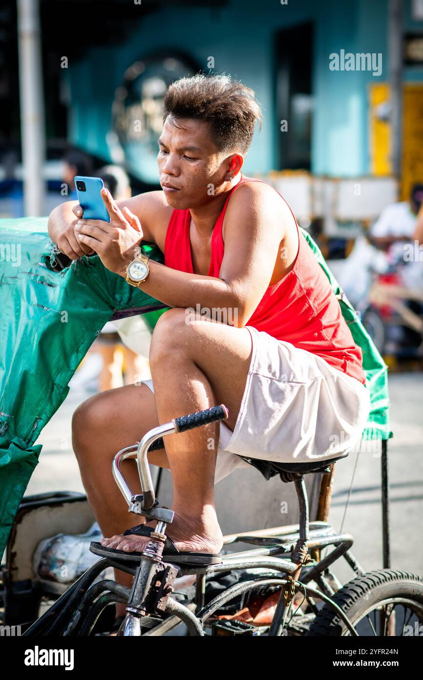 A young filipino man takes a break sat on his trike and checks his ...