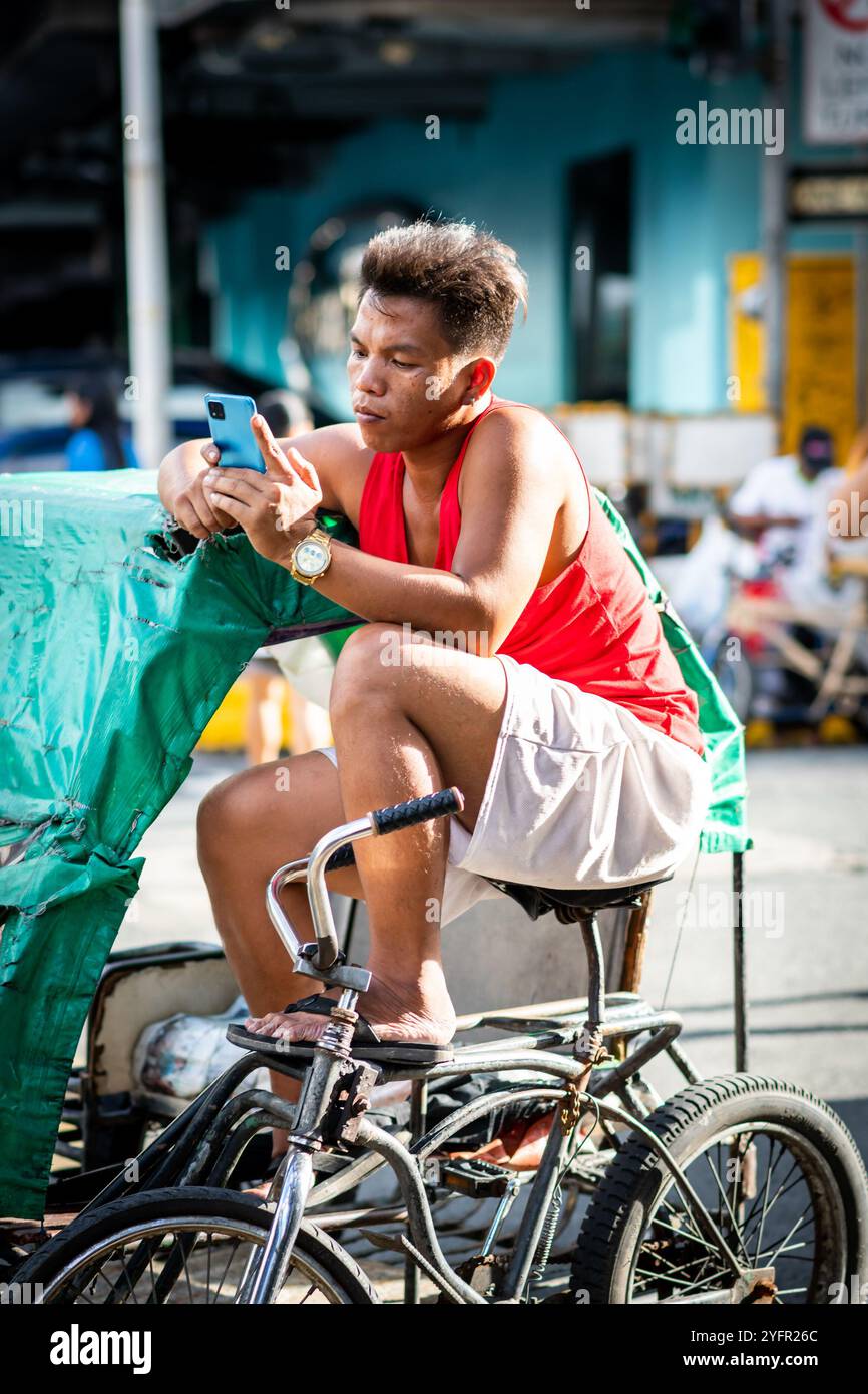 A young filipino man takes a break sat on his trike and checks his ...