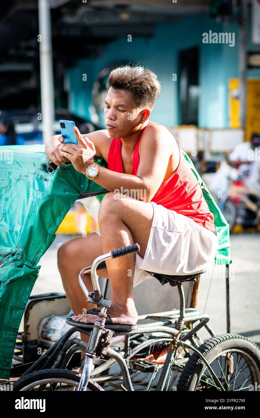 A young filipino man takes a break sat on his trike and checks his ...