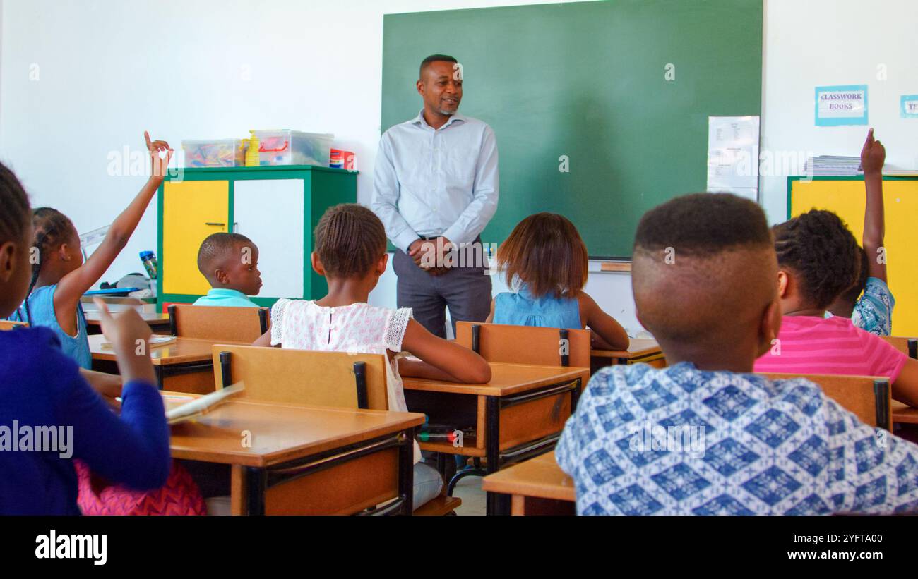 A teacher leads an enthusiastic class of students raising their hands ...