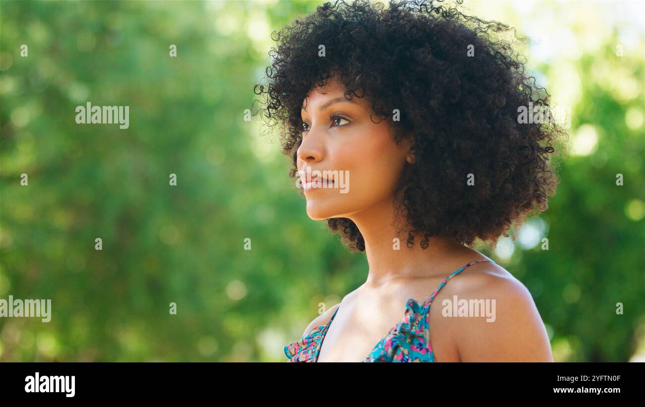 A young woman stands gracefully outdoors, showcasing her curly hair and ...