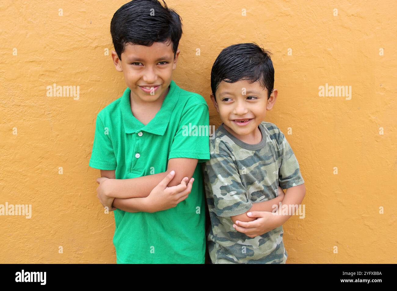 Two dark-haired latin male children standing in front of a yellow wall ...