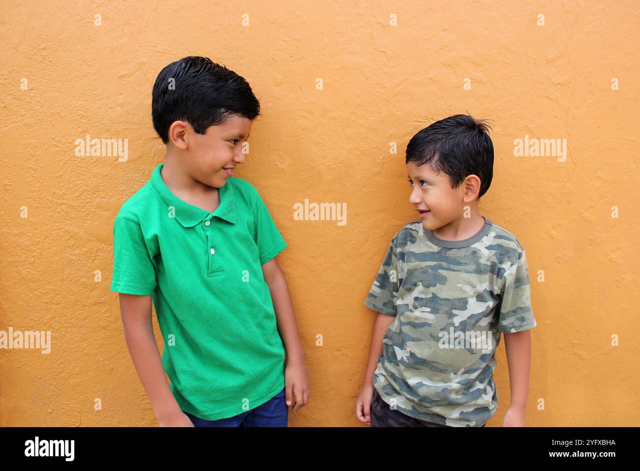 Two dark-haired latin male children standing in front of a yellow wall ...