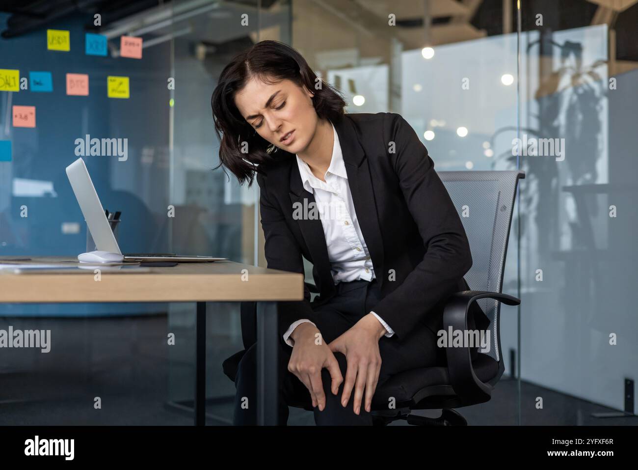 Young businesswoman sitting in office chair, holding knee, depicting ...