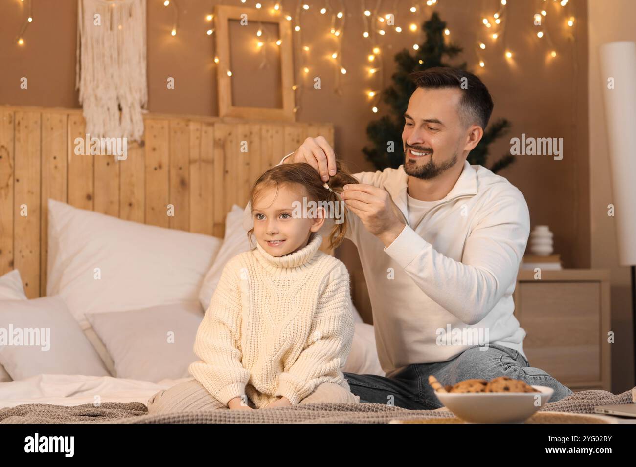 Happy father making ponytails for his daughter on bed in winter evening ...