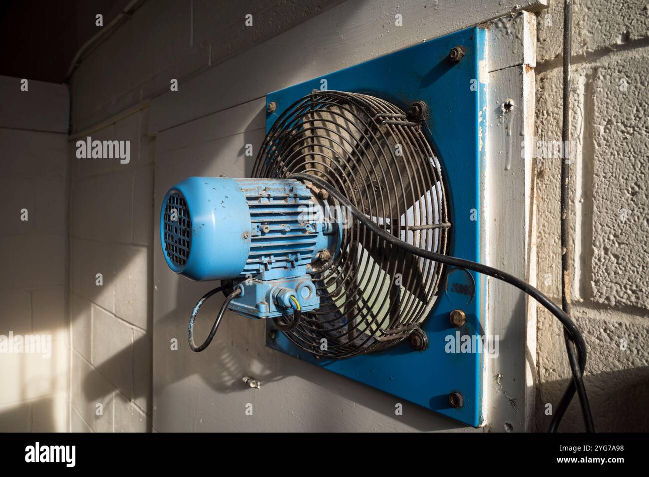 A blue industrial fan mounted on a wall, casting shadows under bright ...