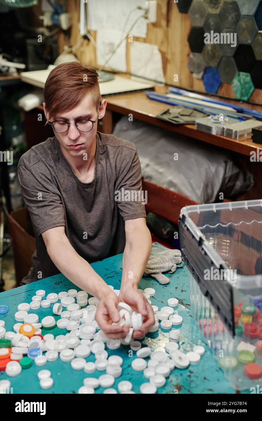 Young male worker of plastic recycling factory sorting polyethylene bottle caps into containers ...