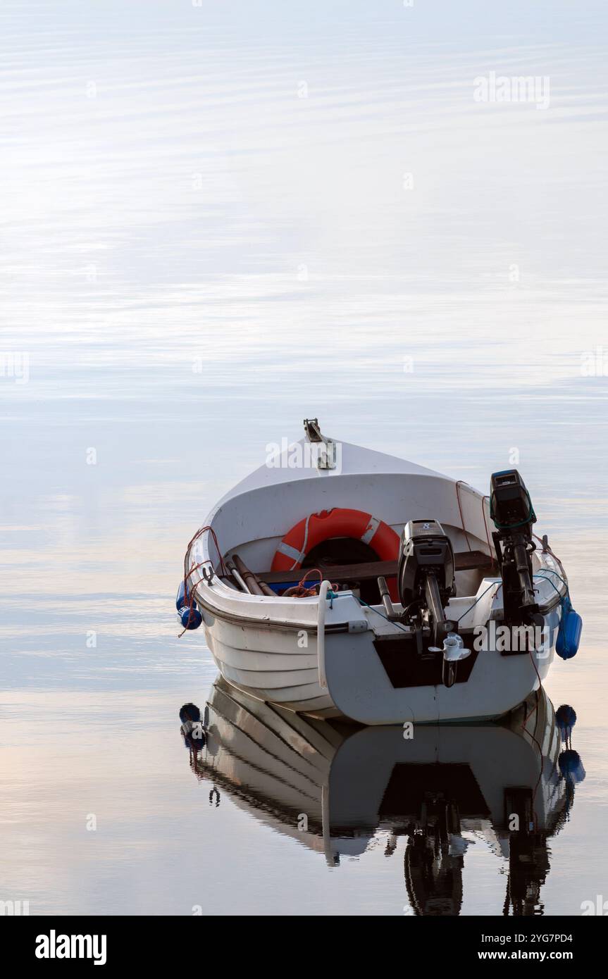 A small boat with two motors sits calmly on the water, its reflection ...